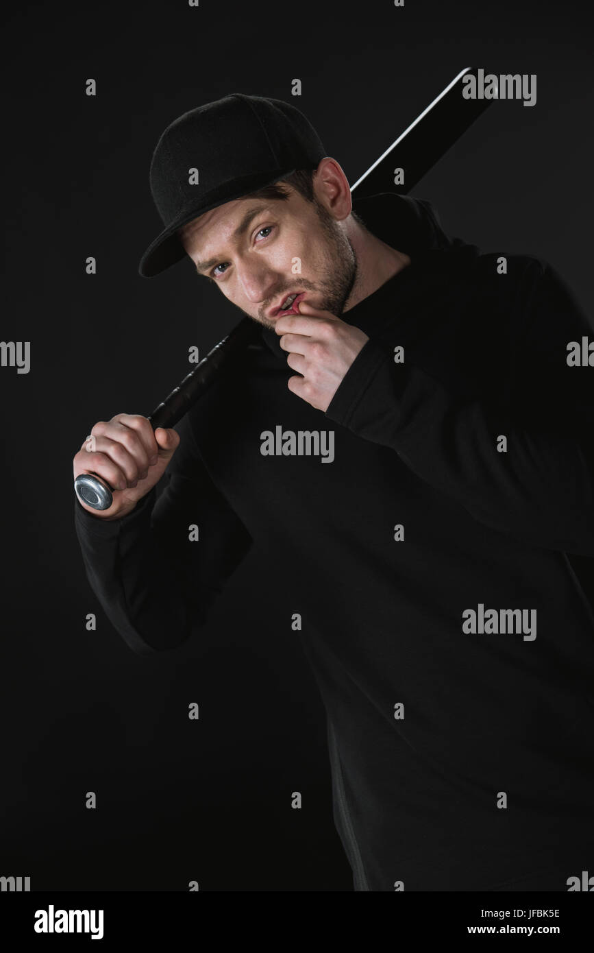 young robber in zoodie and cap with baseball bat isolated on black ...