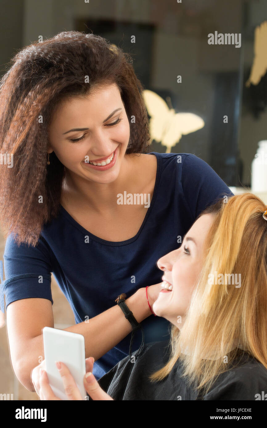 Smiling hairdresser looking on client showing on mobile phone what ...