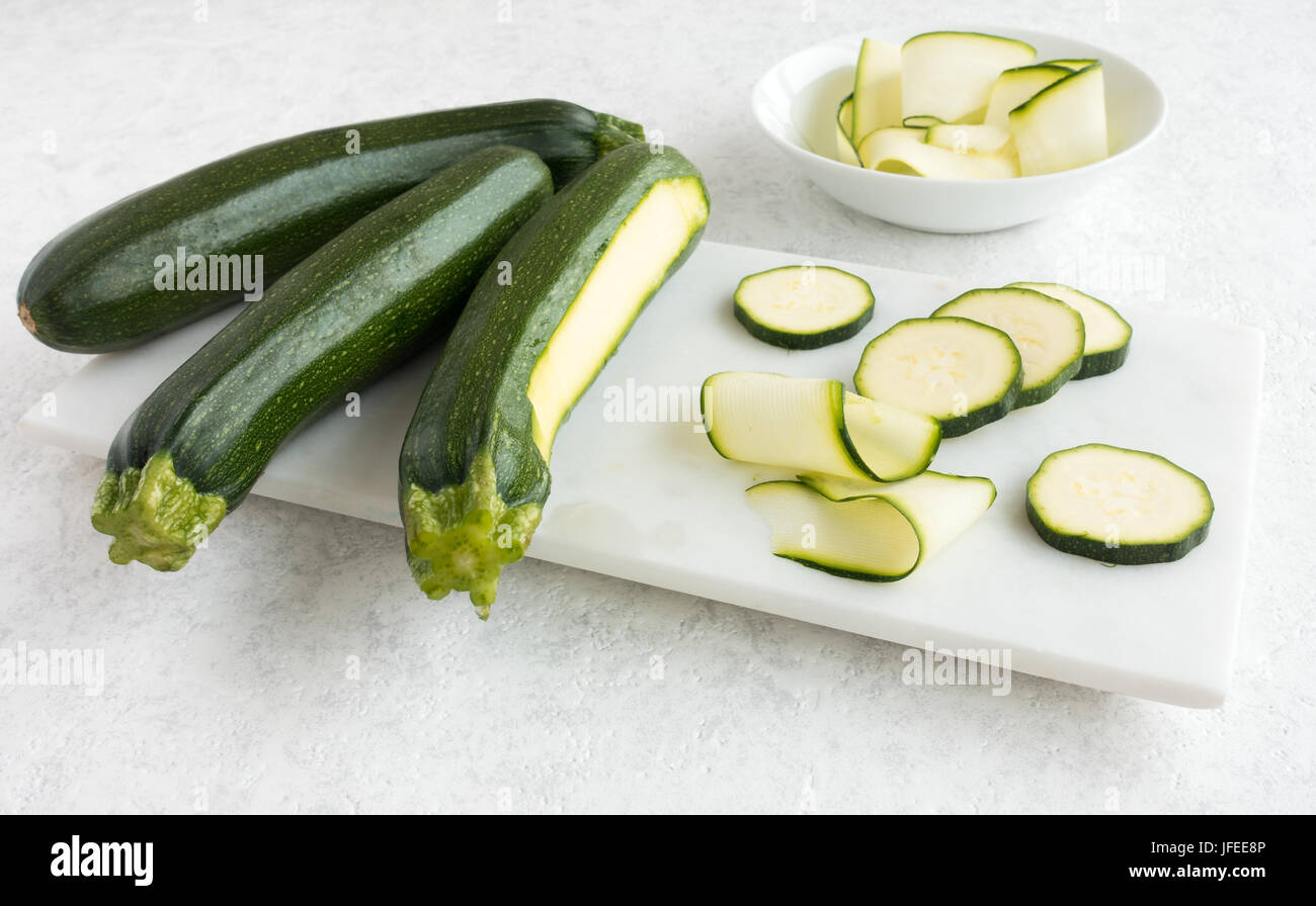 Whole zucchini, curls and slices on white marble chopping board and ...