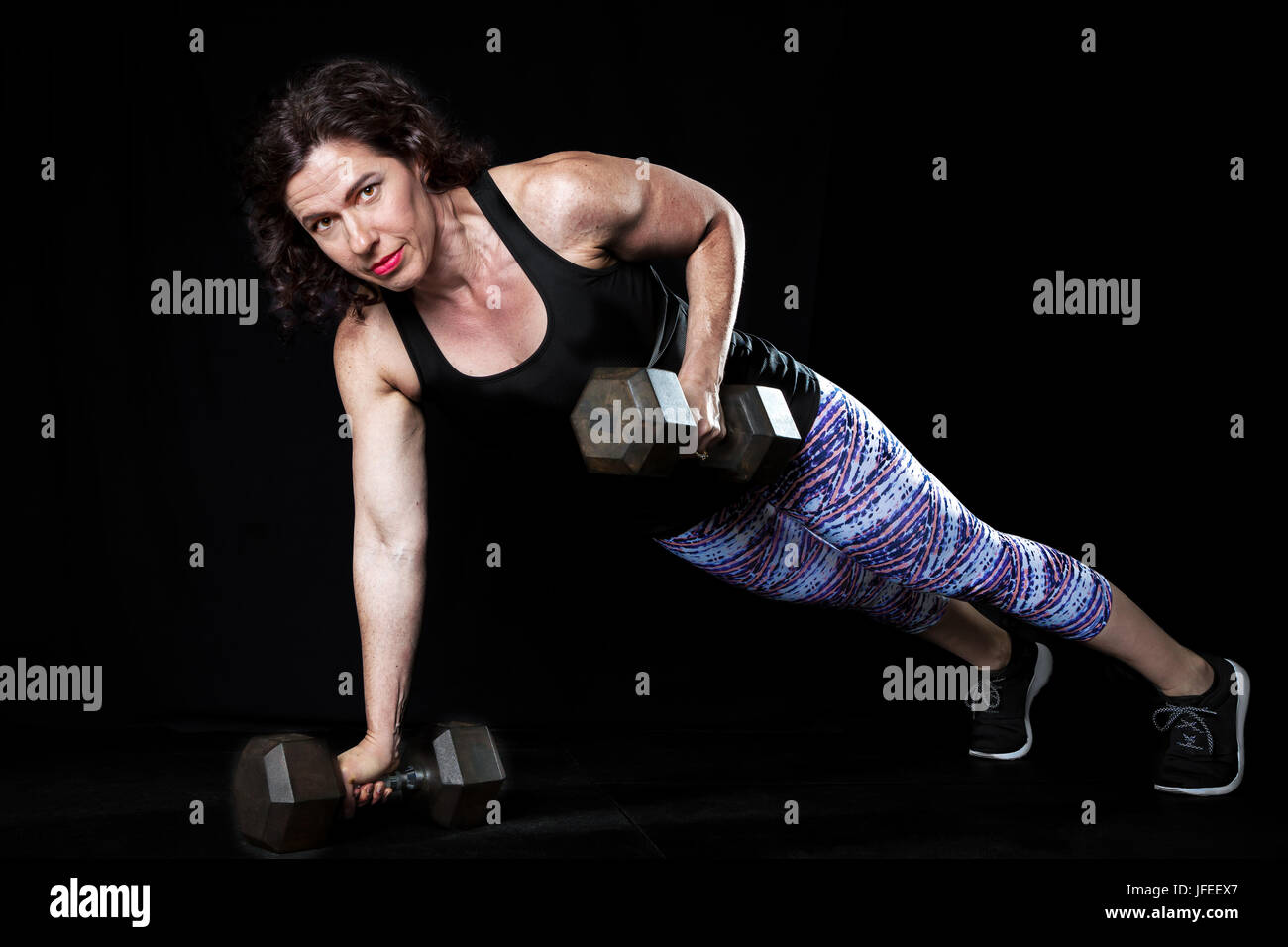 Intense image of a female weightlifter on black, doing a plank dumbell ...