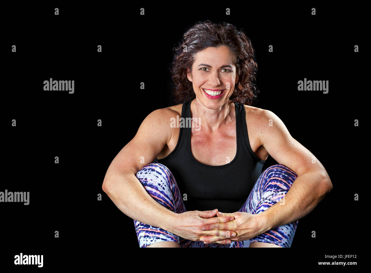 A happy, muscular woman sits with her arms resting on her knees and ...