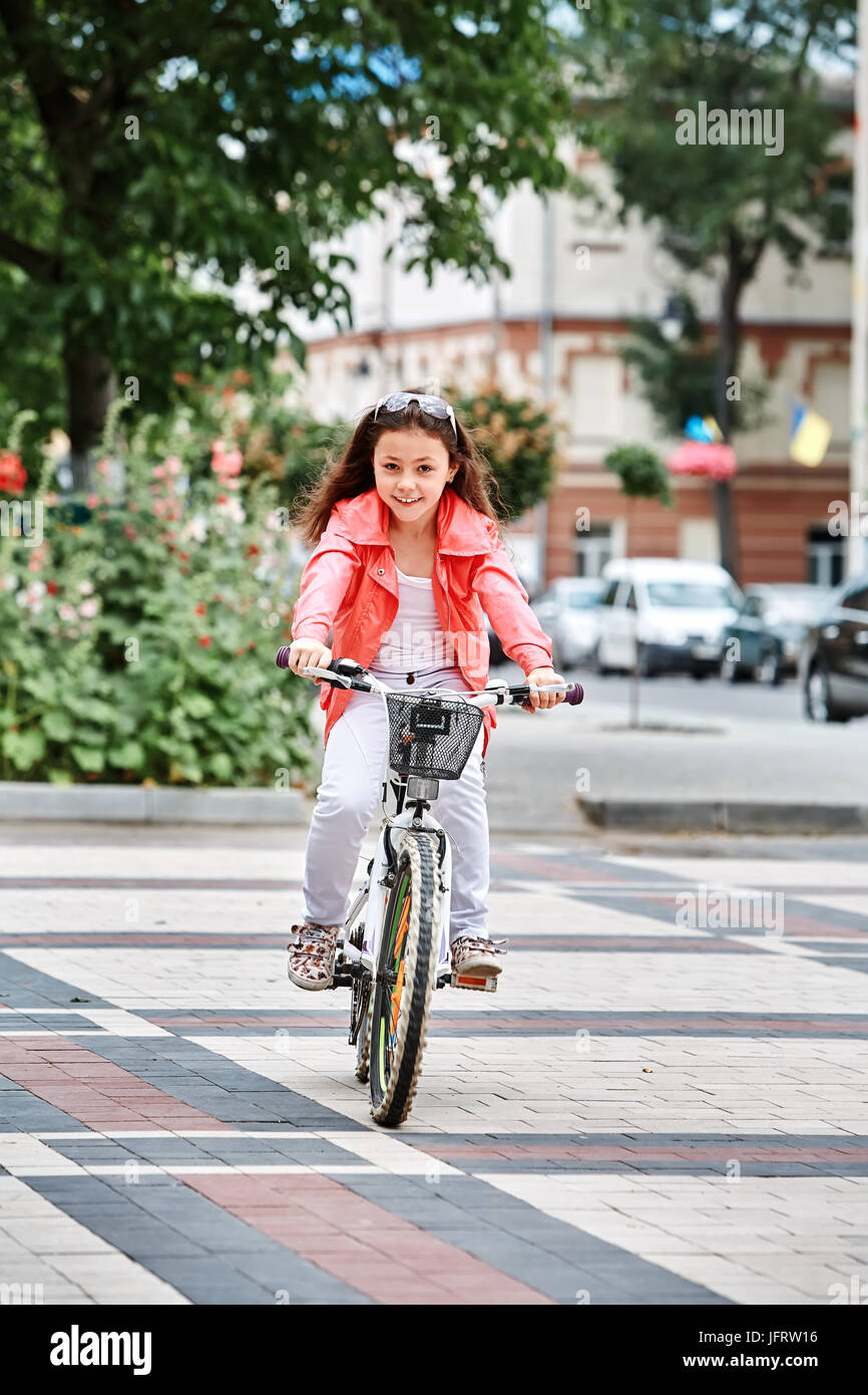 Cute kid girl with long hair in red jacket rides her white bike an city ...