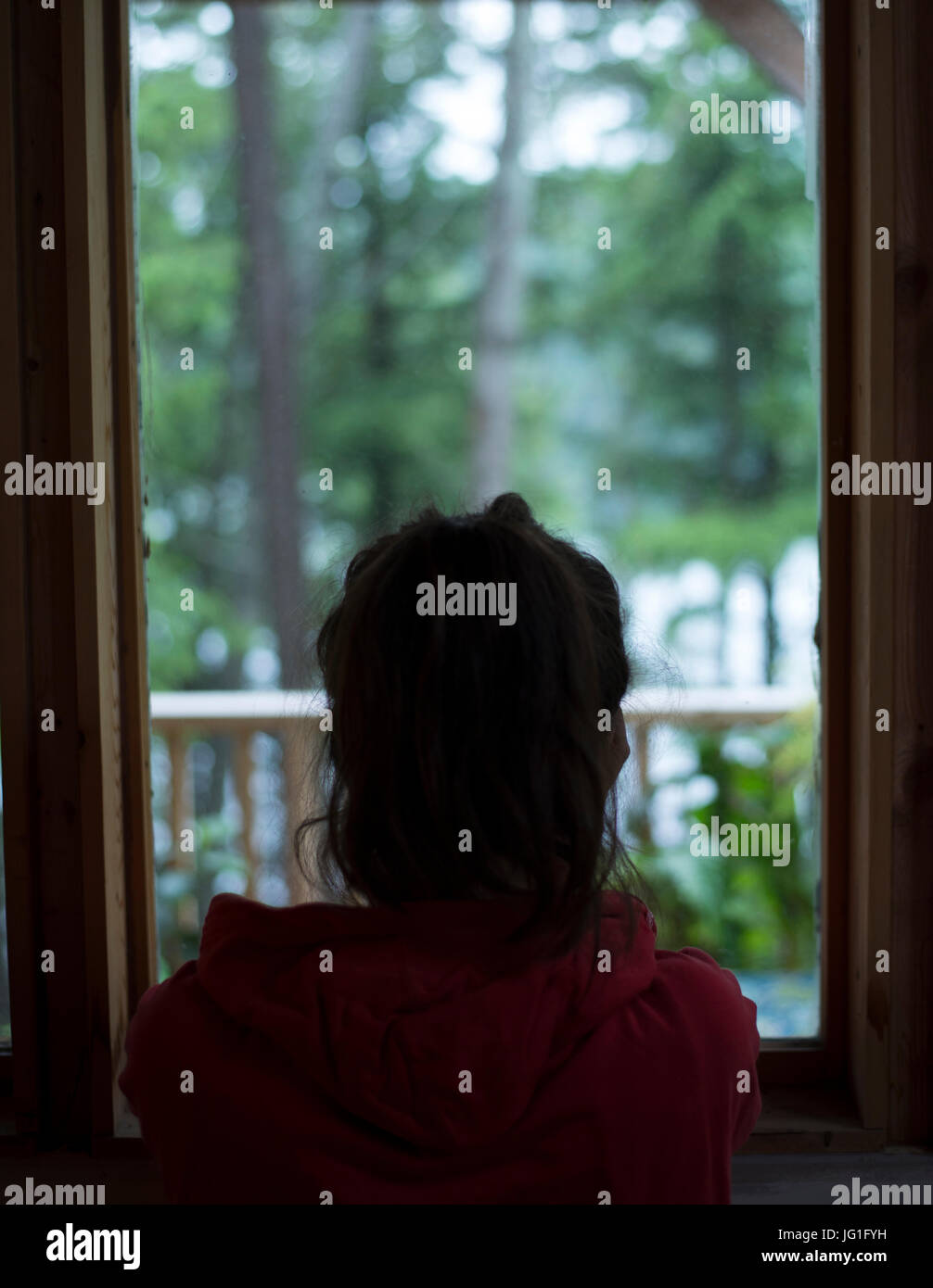 Young brown haired female looking out house window at forest on a rainy ...