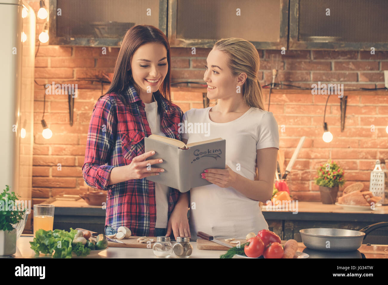Young female friends cooking meal together at home reading receipt book ...