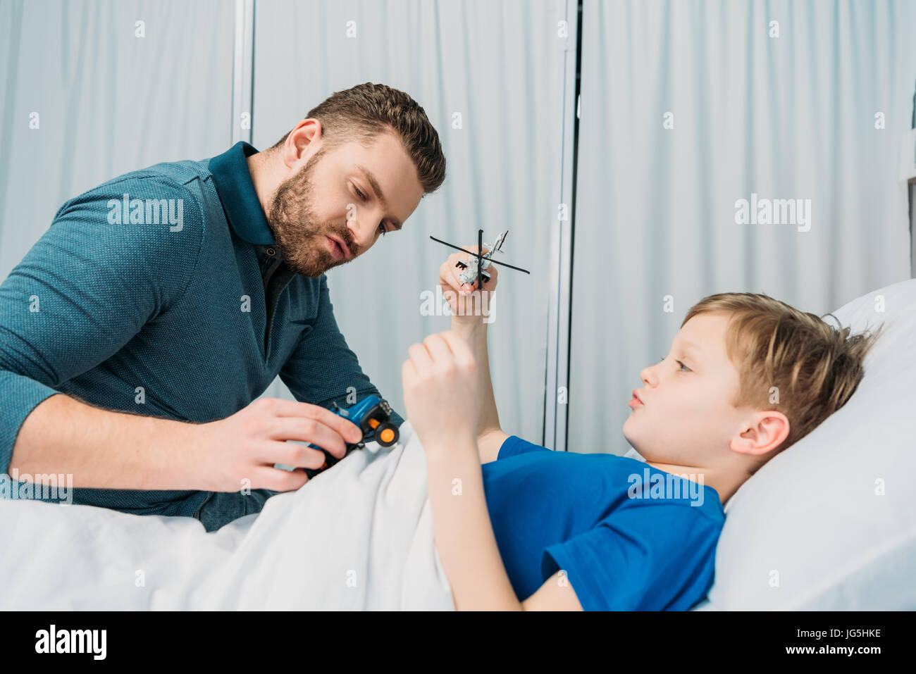 Smiling father playing with sick little boy lying in hospital bed, dad ...
