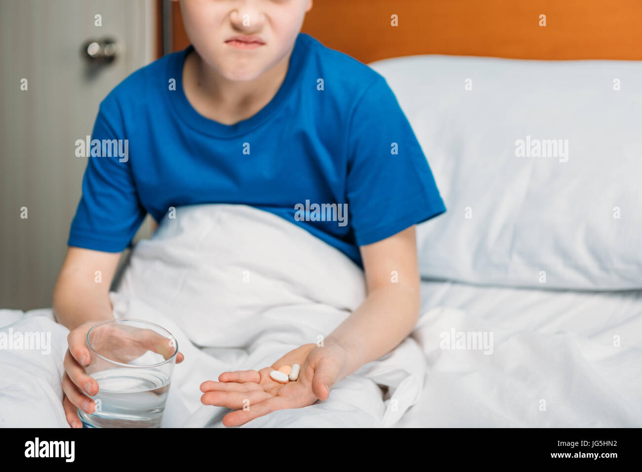 unsatisfied boy holding glass of water and medicines in hospital bed ...