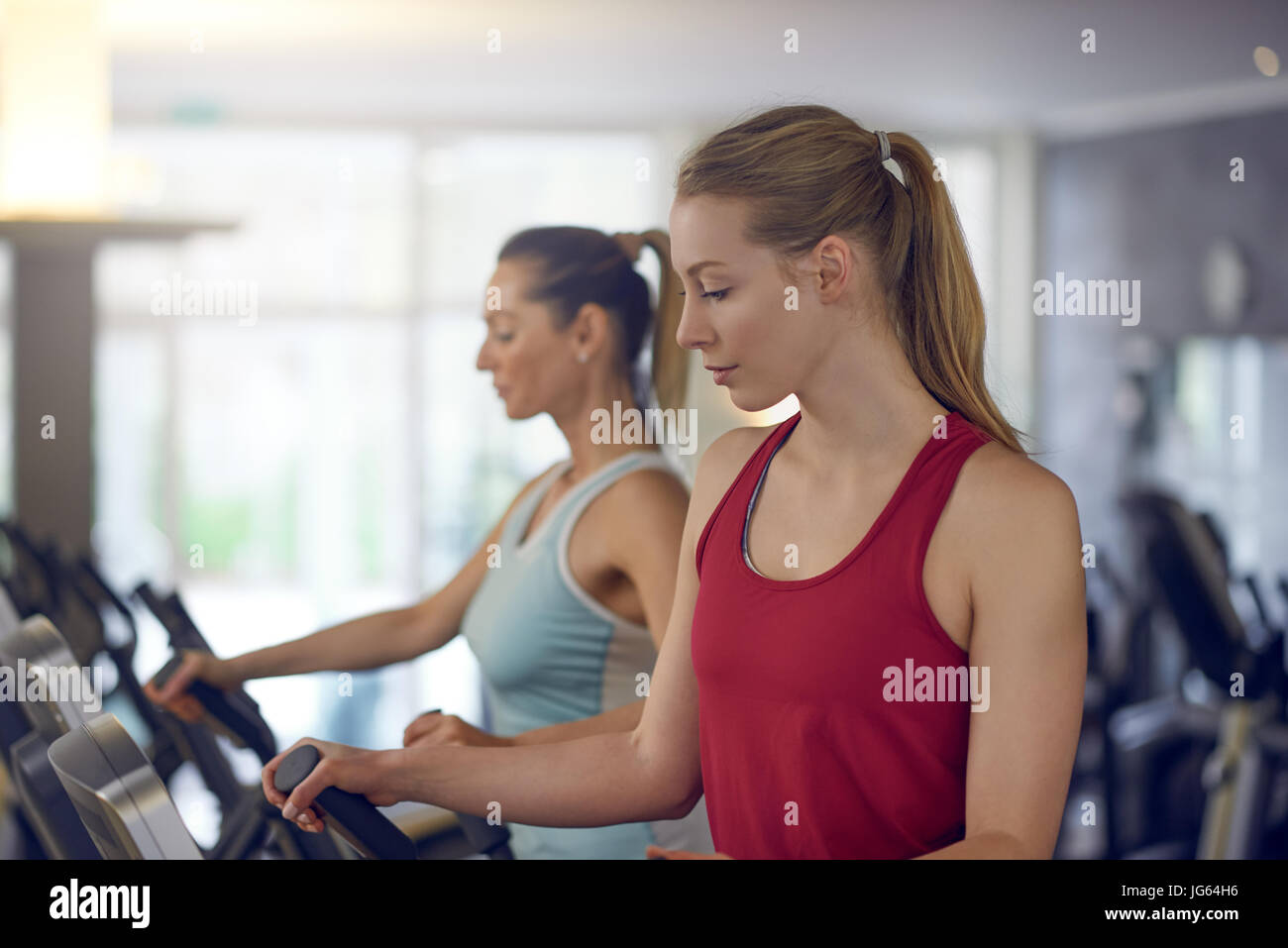 Two healthy women working out in a gym on exercise machines with focus ...