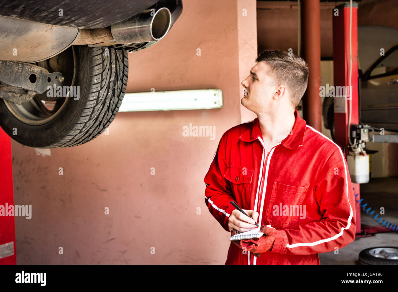 Male auto mechanic standing making notes underneath a lifted car near ...
