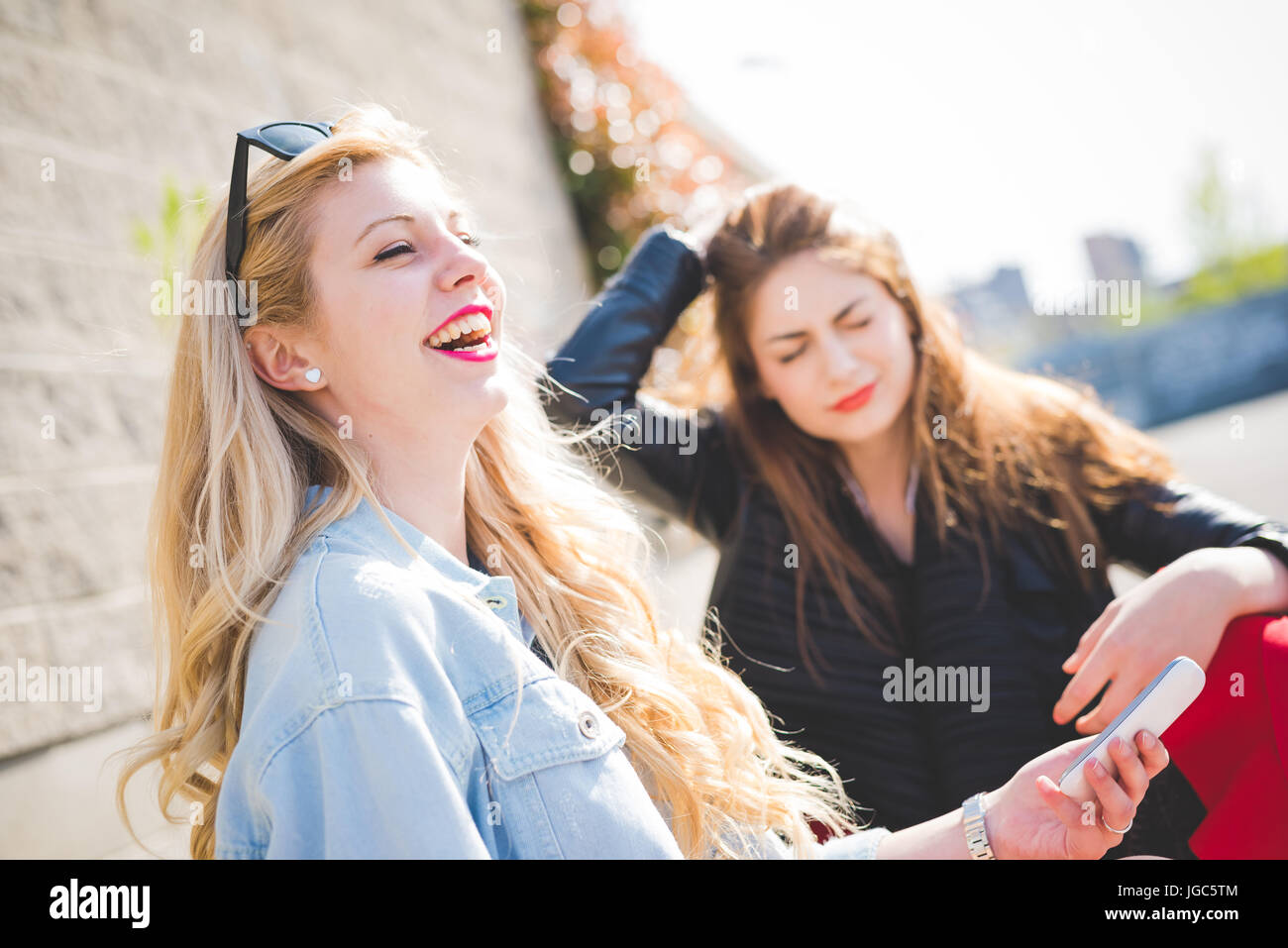 Two beautiful young women sitting outdoor using smart phone interacting ...