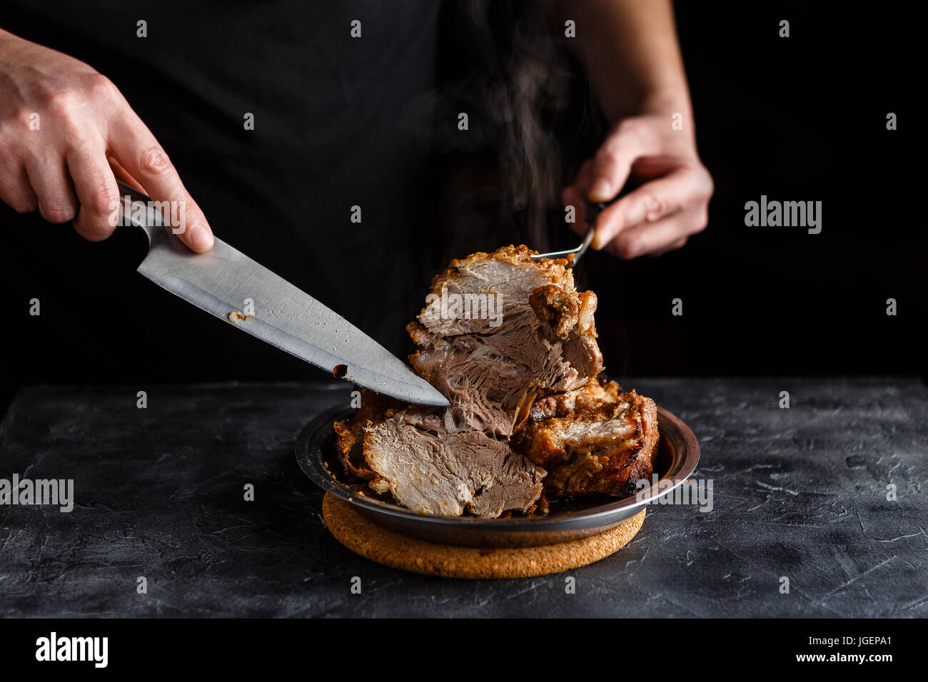 Man cuts roasted pieces of pork shoulder blade in metal plate on dark ...
