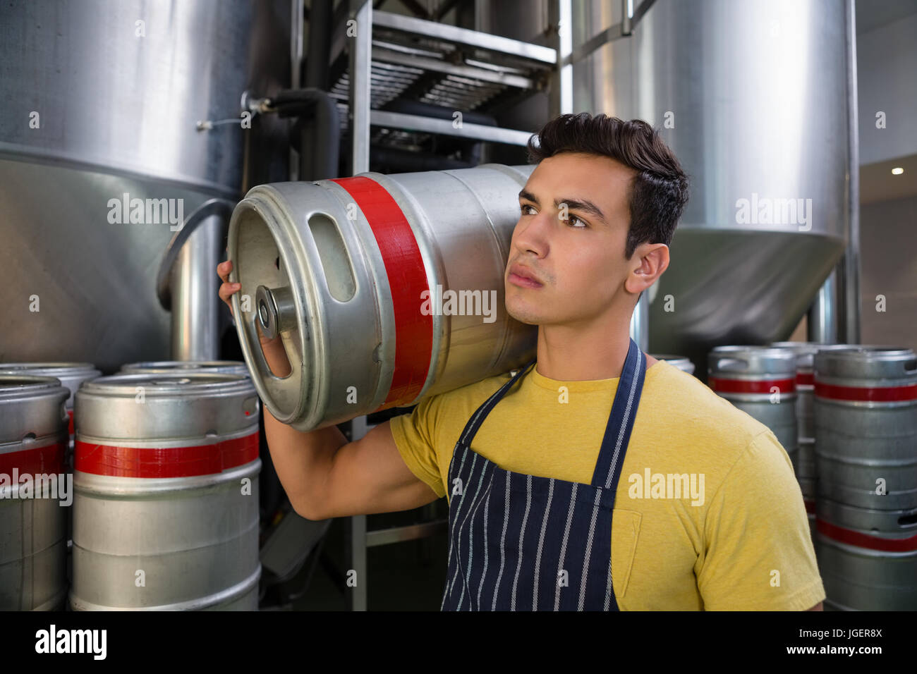 Worker looking away while carrying keg by storage tanks at brewery ...