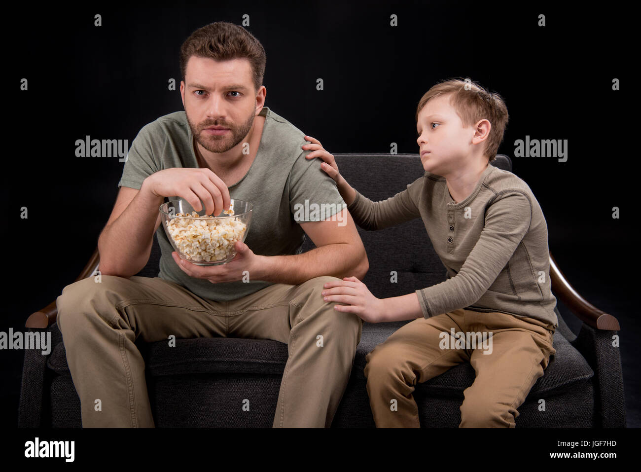 Little boy trying to talk with father eating popcorn and watching tv on ...