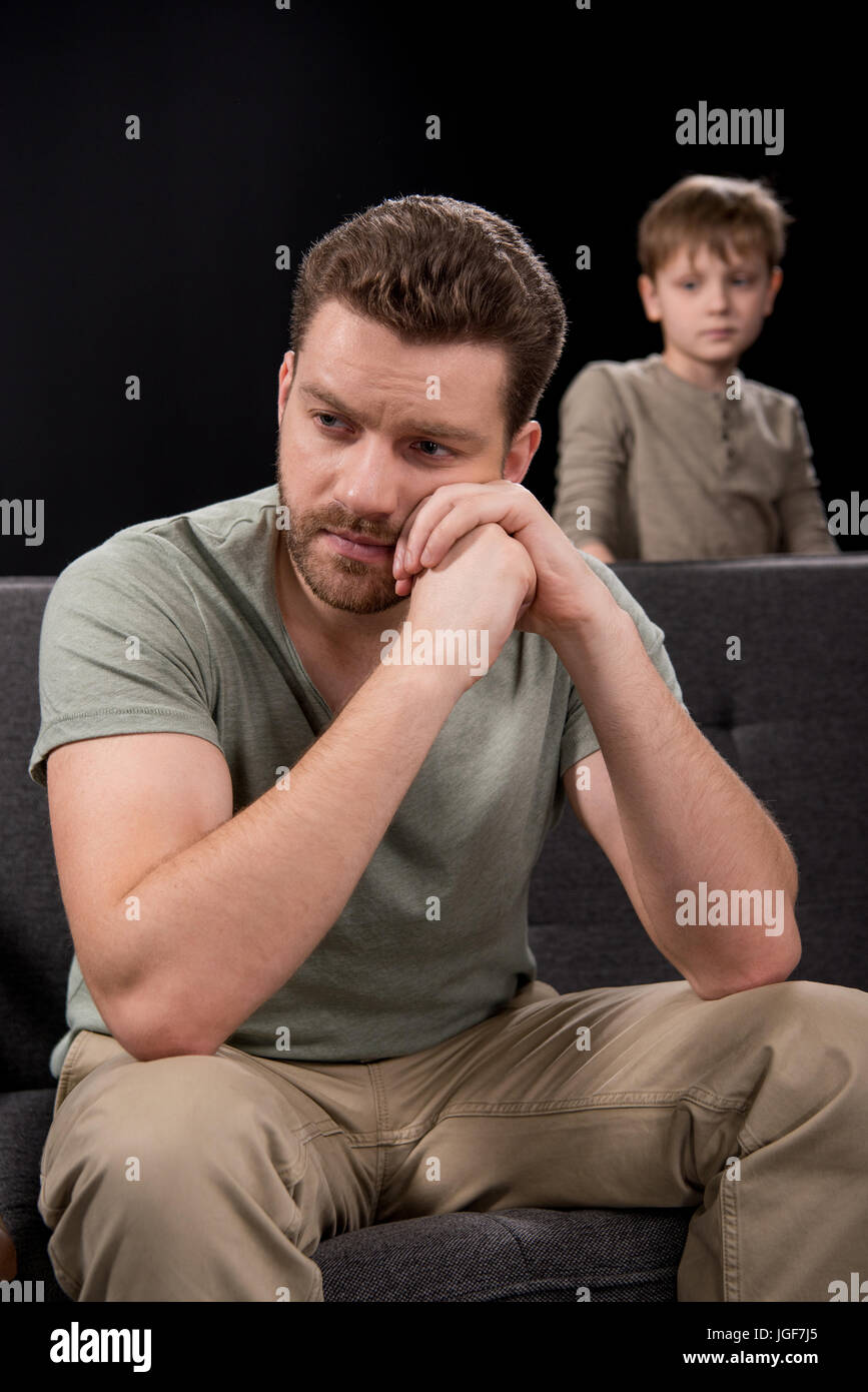 Upset little boy looking at pensive father sitting on sofa and looking ...