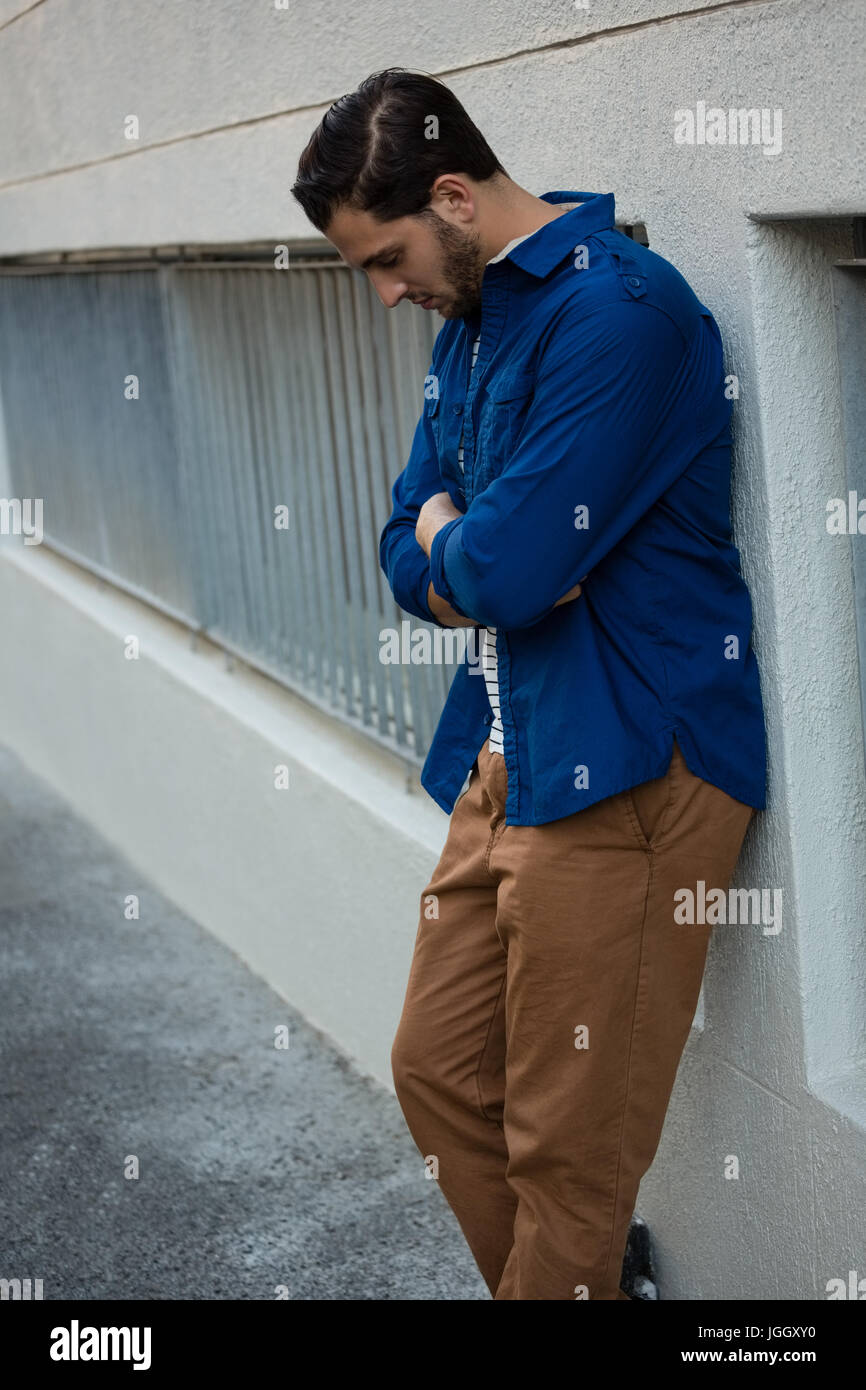Thoughtful man with arms crossed looking down while leaning on wall ...