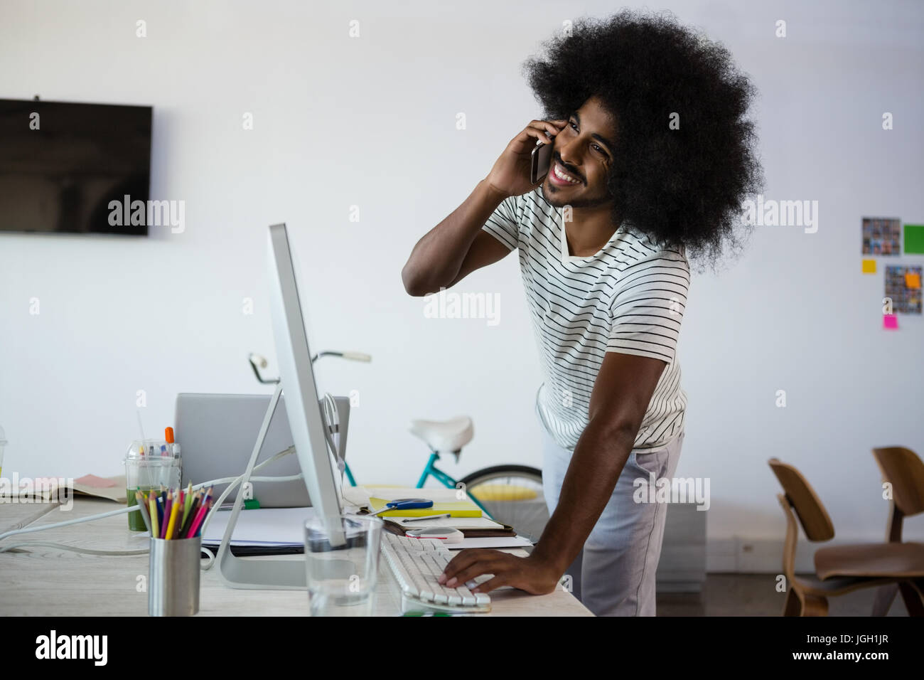 Smiling young man talking on phone while using computer at desk in ...