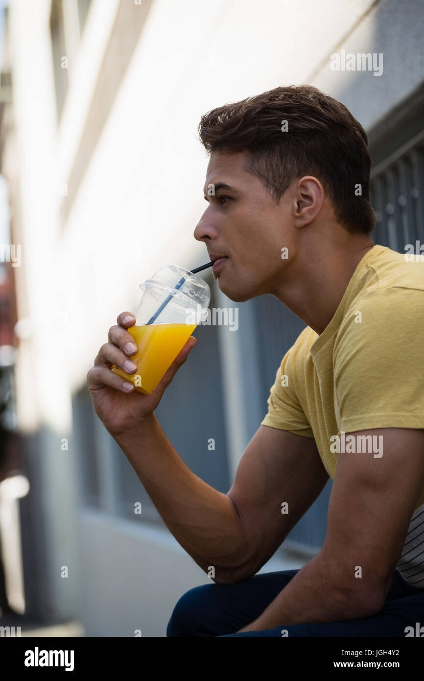 Side view of young man drinking juice while sitting by wall in city ...