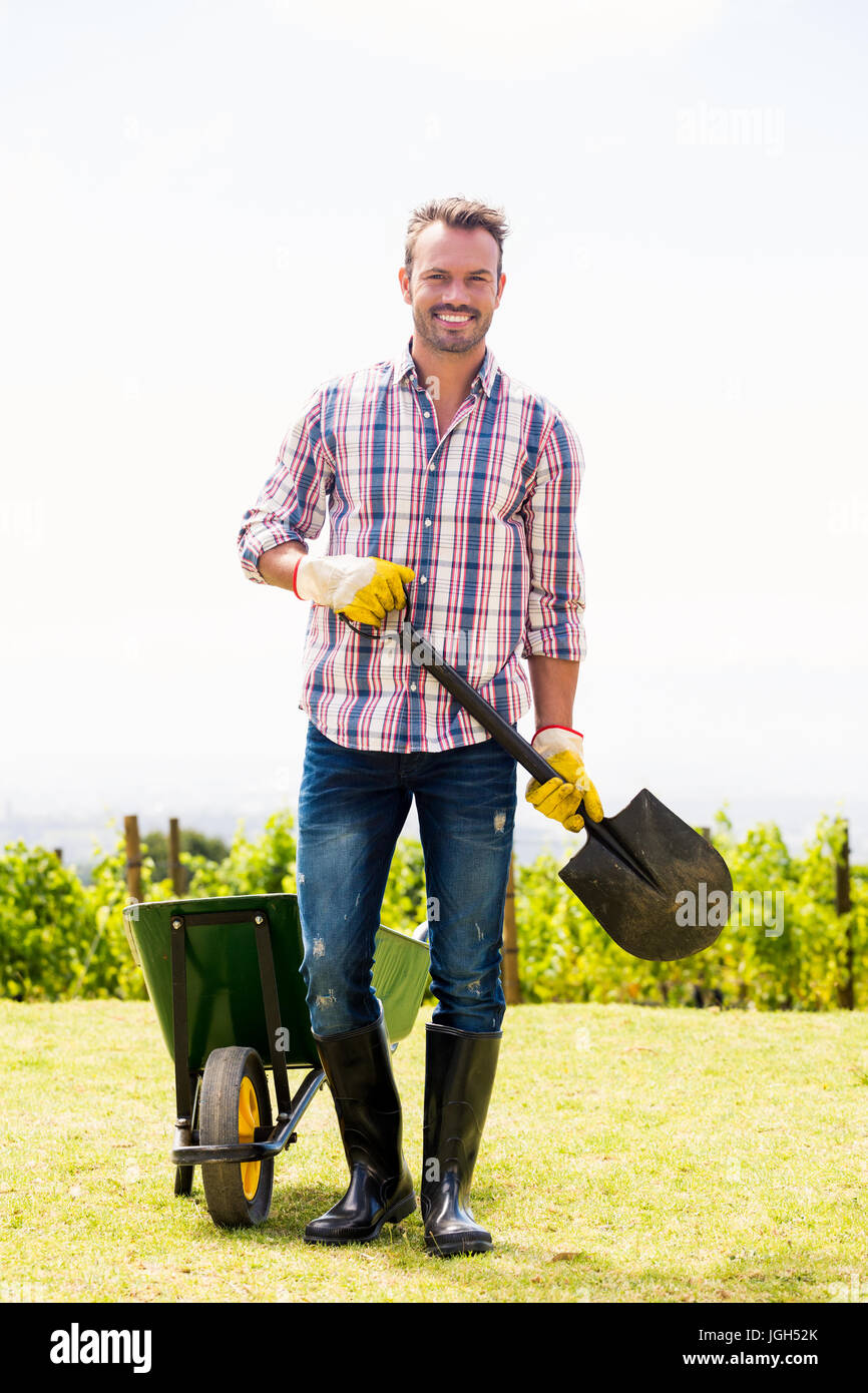 Portrait of young man holding shovel by wheelbarrow against sky at lawn ...