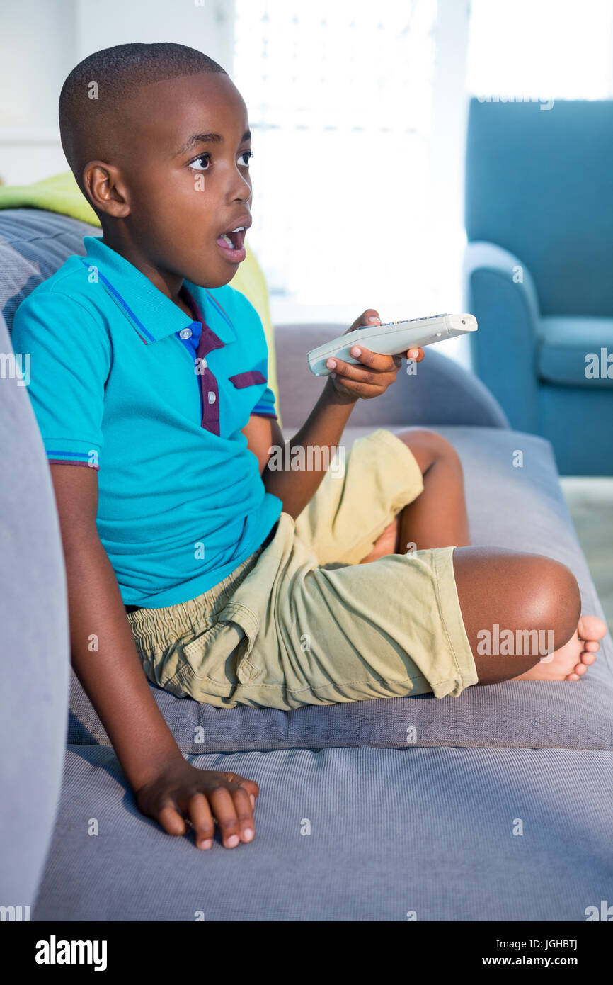 Surprised boy holding remote control while sitting on sofa in living ...