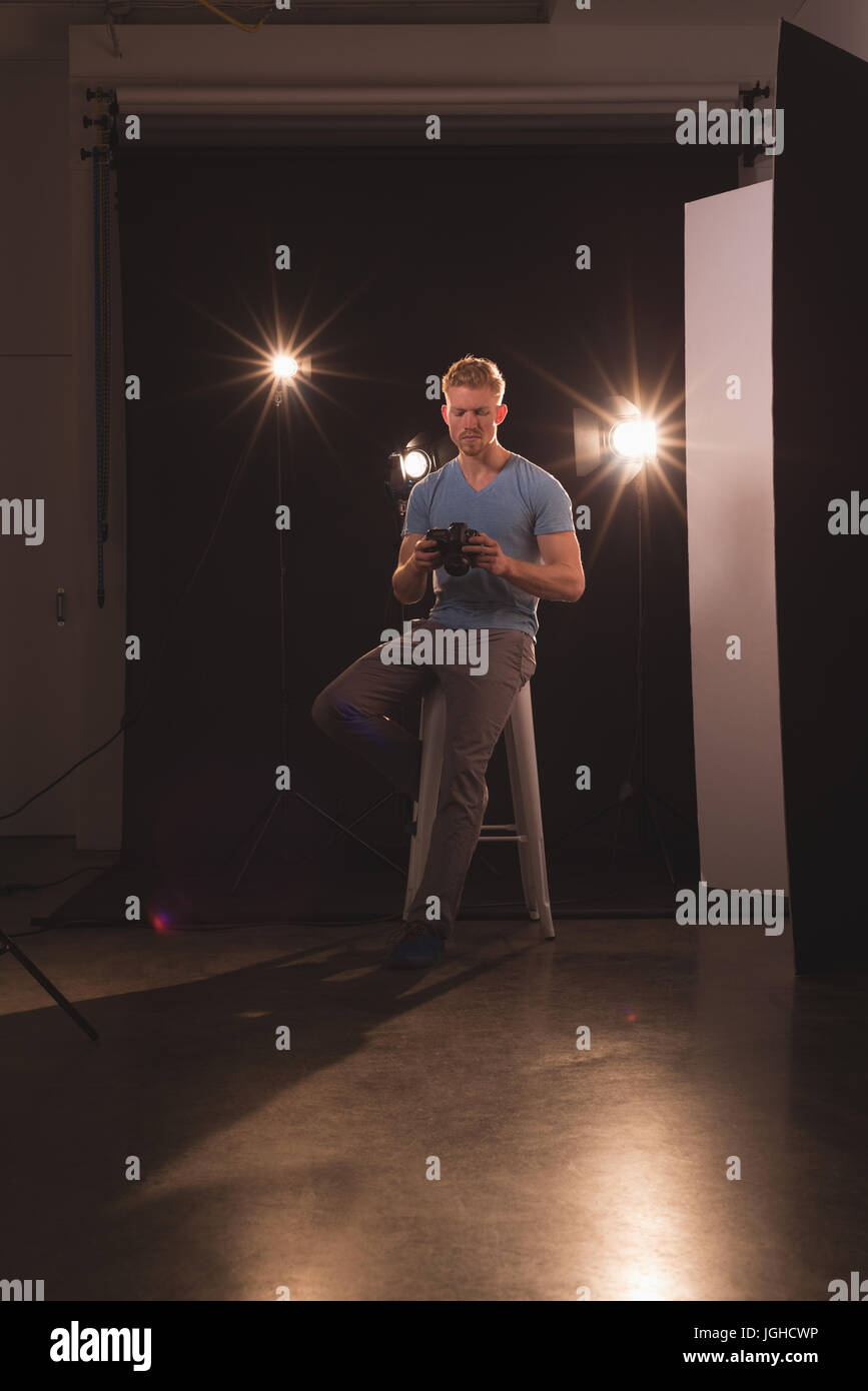Full length of man adjusting camera while sitting on stool against ...