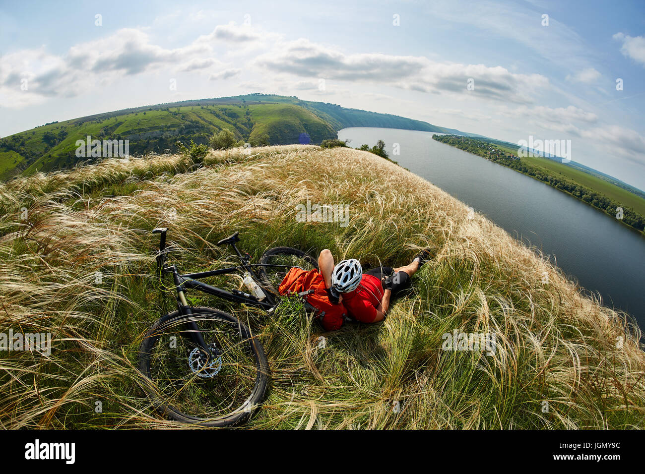 The attractive cyclist sitting on the grass on the meadow with mountain ...