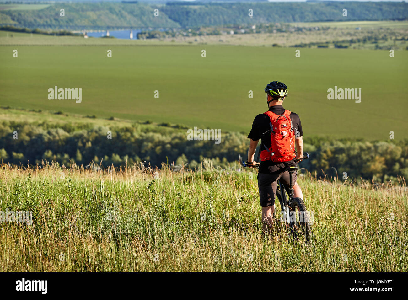 Rear view of the sportsman with his mountain bike stands on the meadow ...