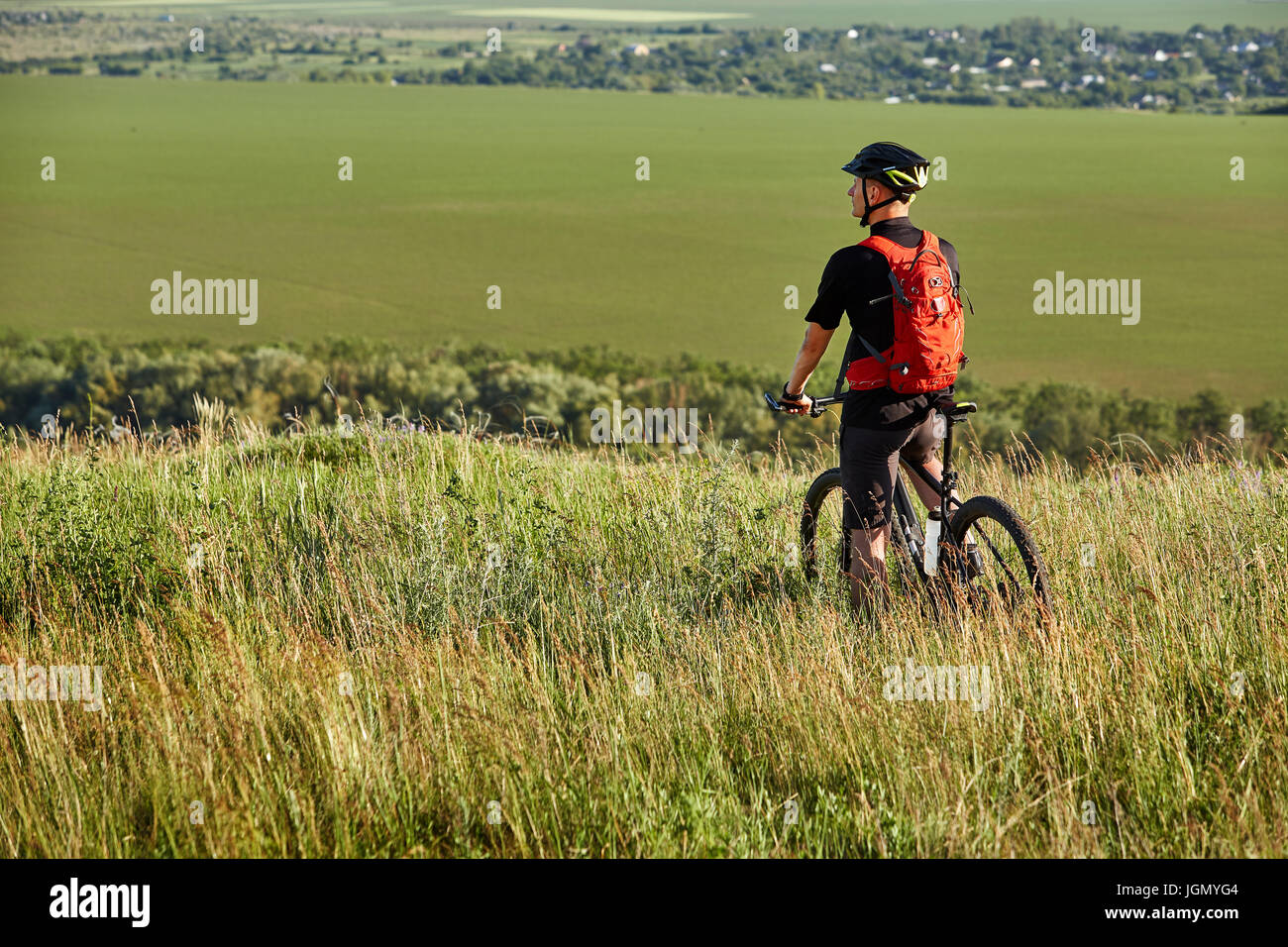 Rear view of the sportsman with his mountain bike stands on the meadow ...