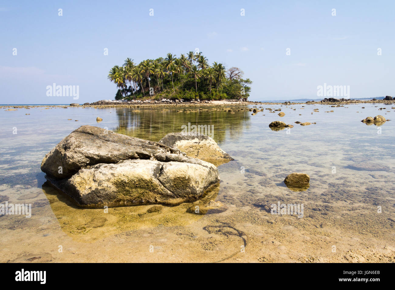 Foreground rocks in clear water with a small island with palm trees ...