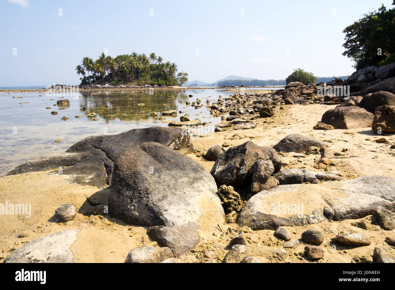 Foreground rocks in clear water with a small island with palm trees ...
