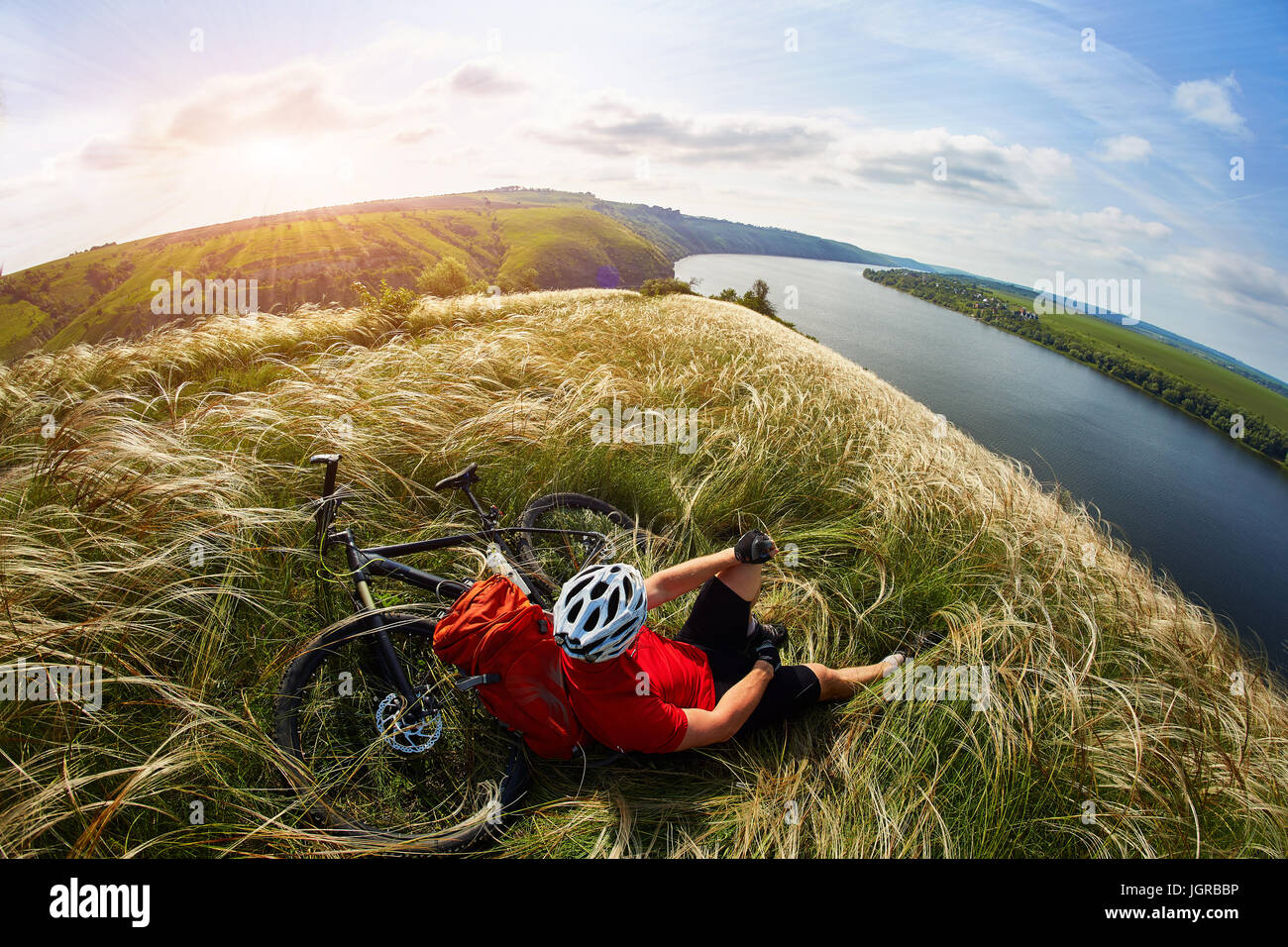 The attractive cyclist sitting on the grass on the meadow with mountain ...