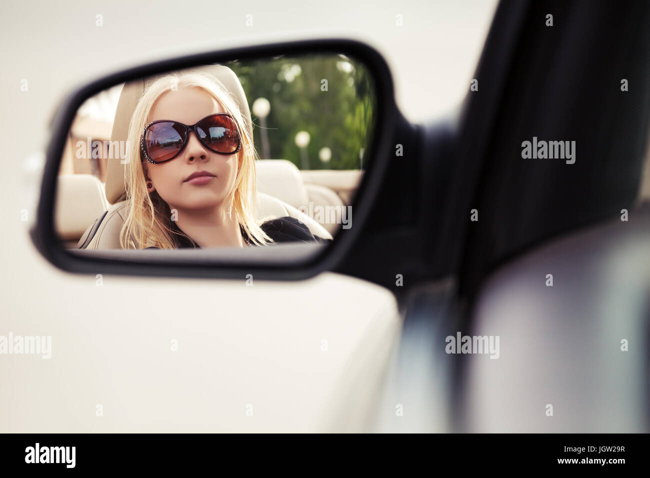 Blond fashion woman in sunglasses looking in the car rear view mirror ...