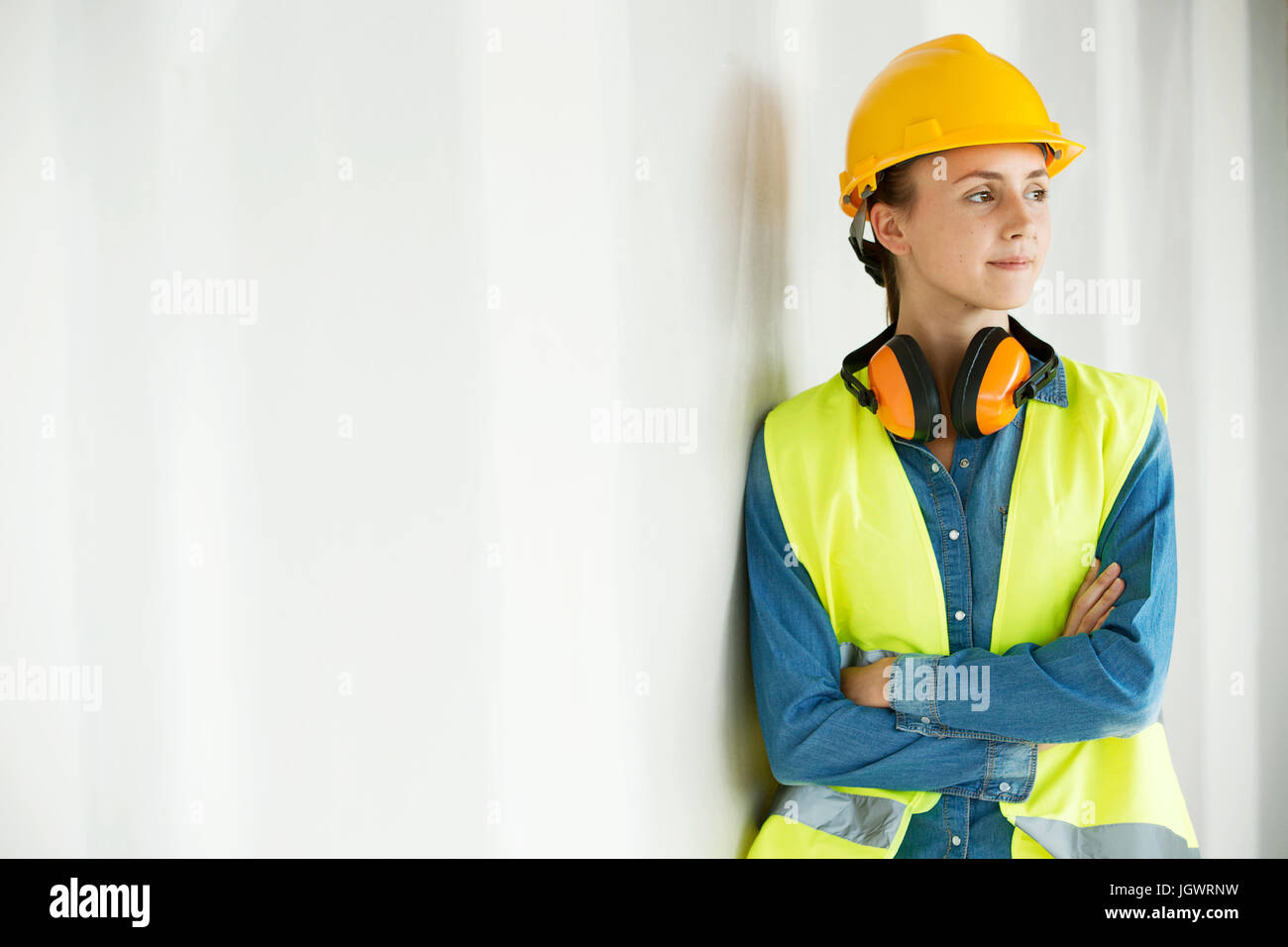Young woman, wearing hi vis vest and hard hat, leaning against wall ...