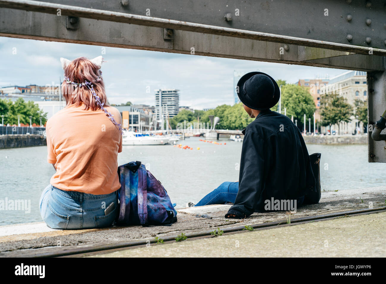Two young people sitting on the docks looking at a view of the harbour ...