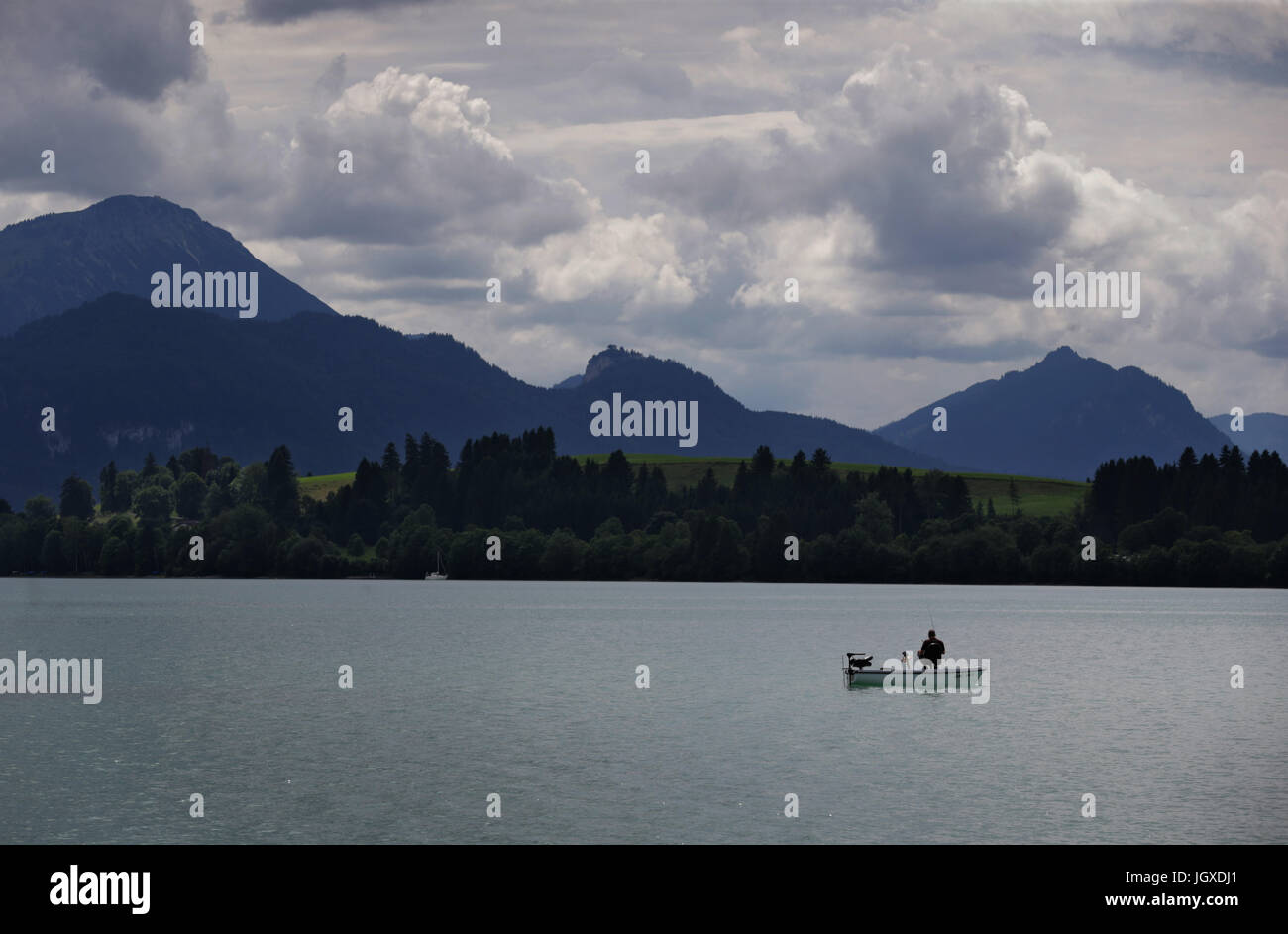 A man fishes on a boat at the lake Forggensee while clouds move across ...