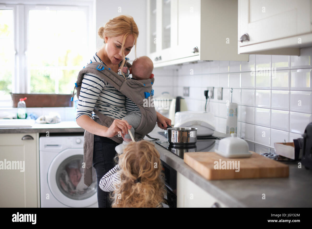 Woman cooking in kitchen, baby strapped to body in sling, daughter ...