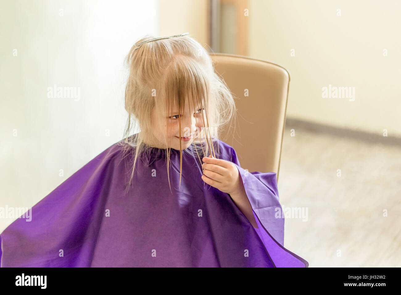 Little cute blond girl smiles and looks at hairdresser during haircut ...