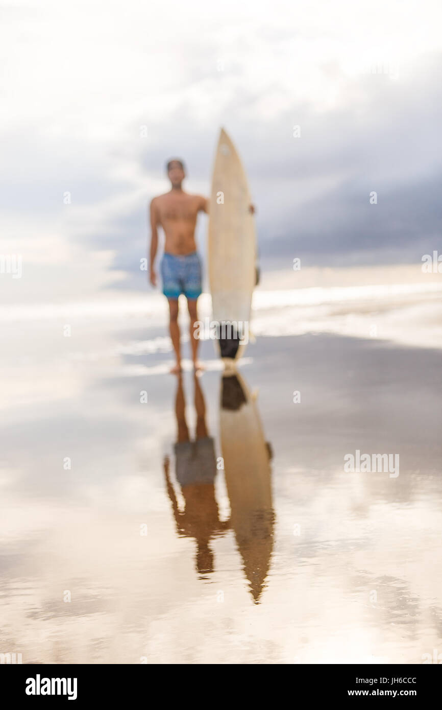 Out of focus, blurry image of handsome man with surfboard stand at sand ...