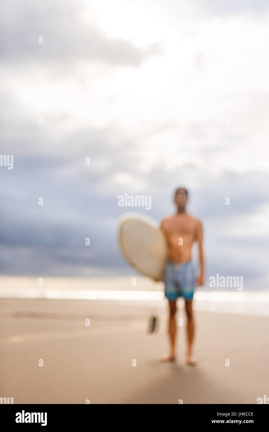 Out of focus, blurry image of handsome boy with surfboard stand at sand ...