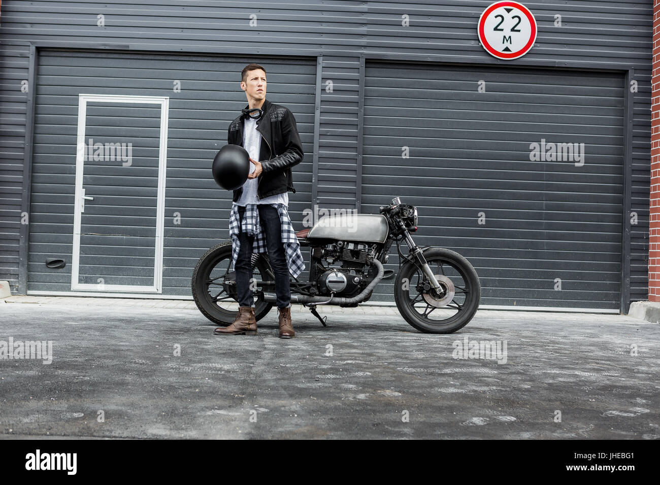 Handsome rider biker man hold his black helmet near industrial gates ...