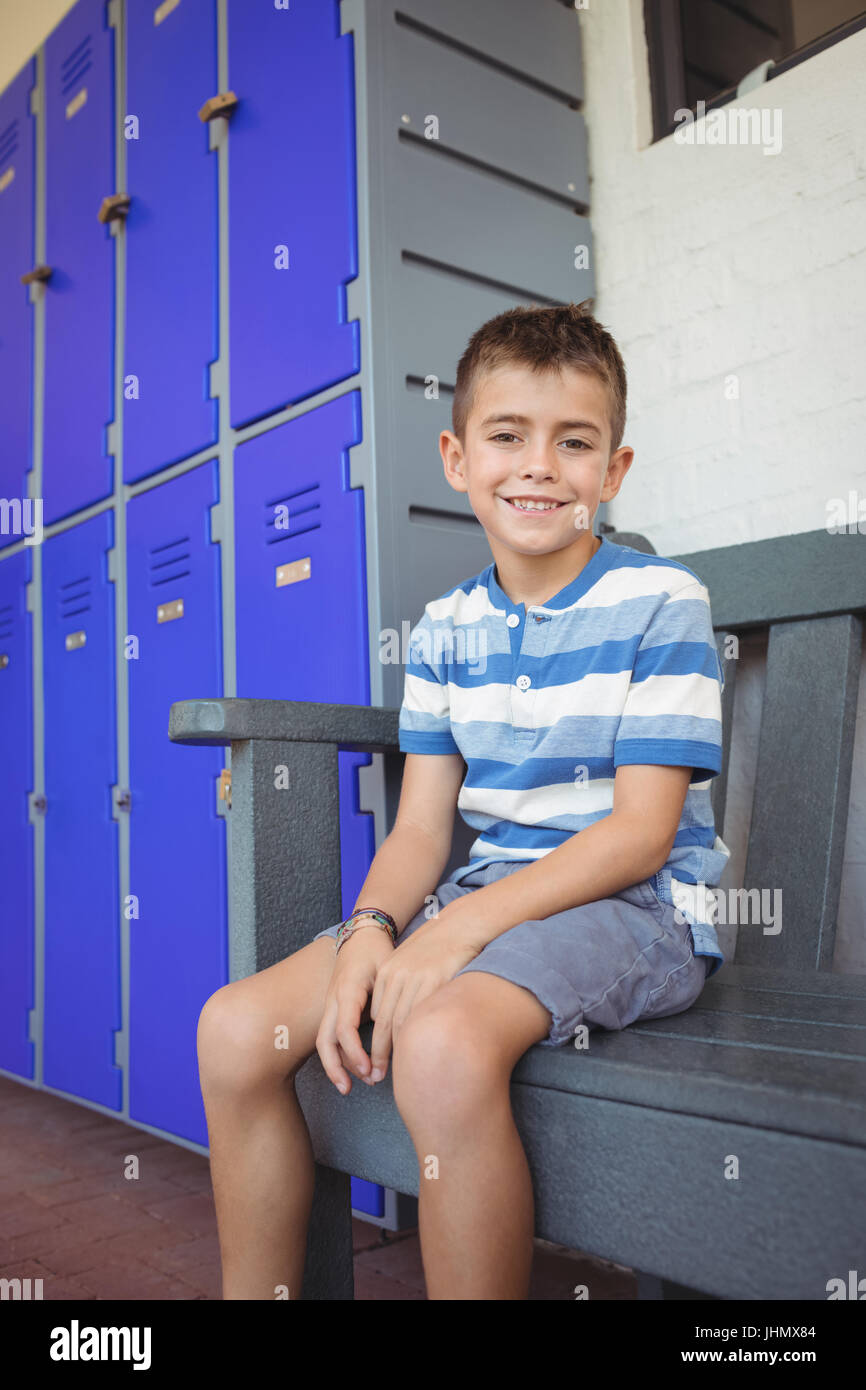 Portrait of smiling boy sitting on bench by lockers in corridor at ...