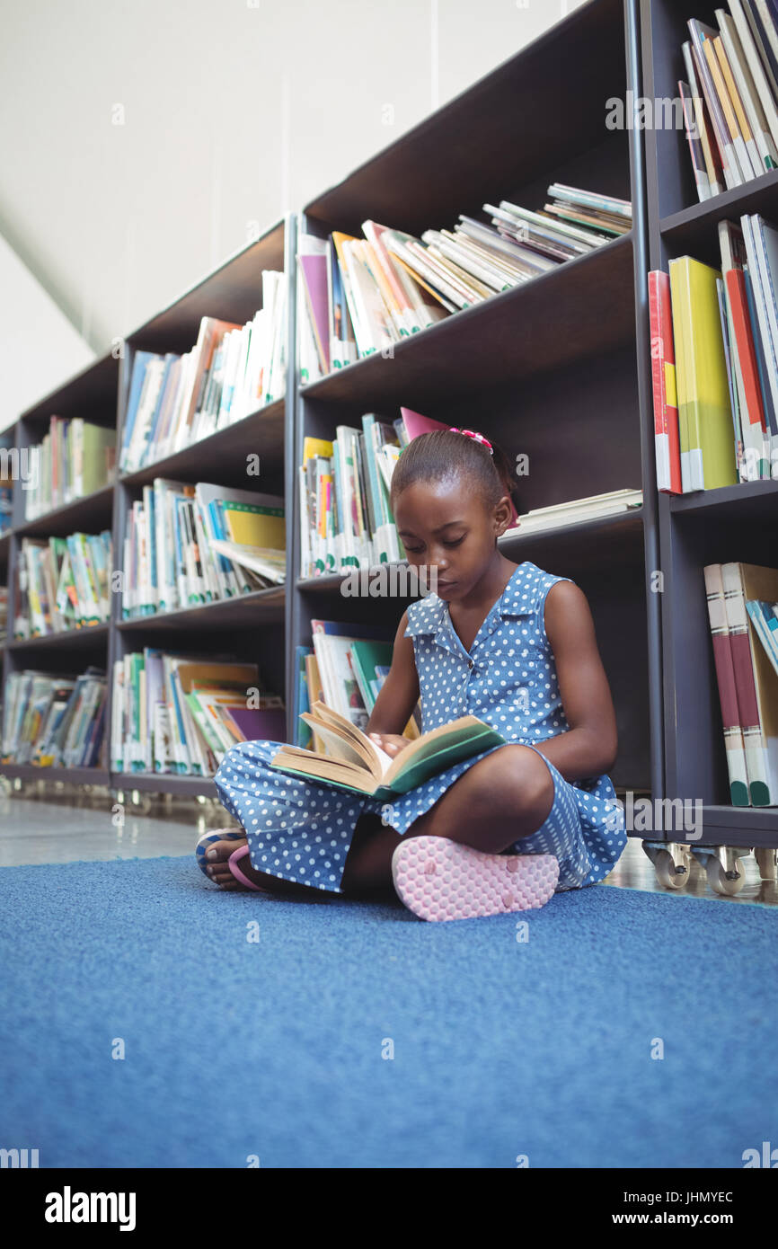 Girl reading book while sitting by bookshelf in library Stock Photo - Alamy