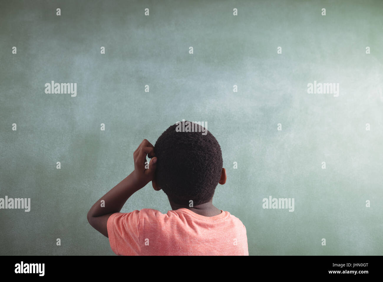 Rear view of thoughtful boy standing against greenboard in classroom ...