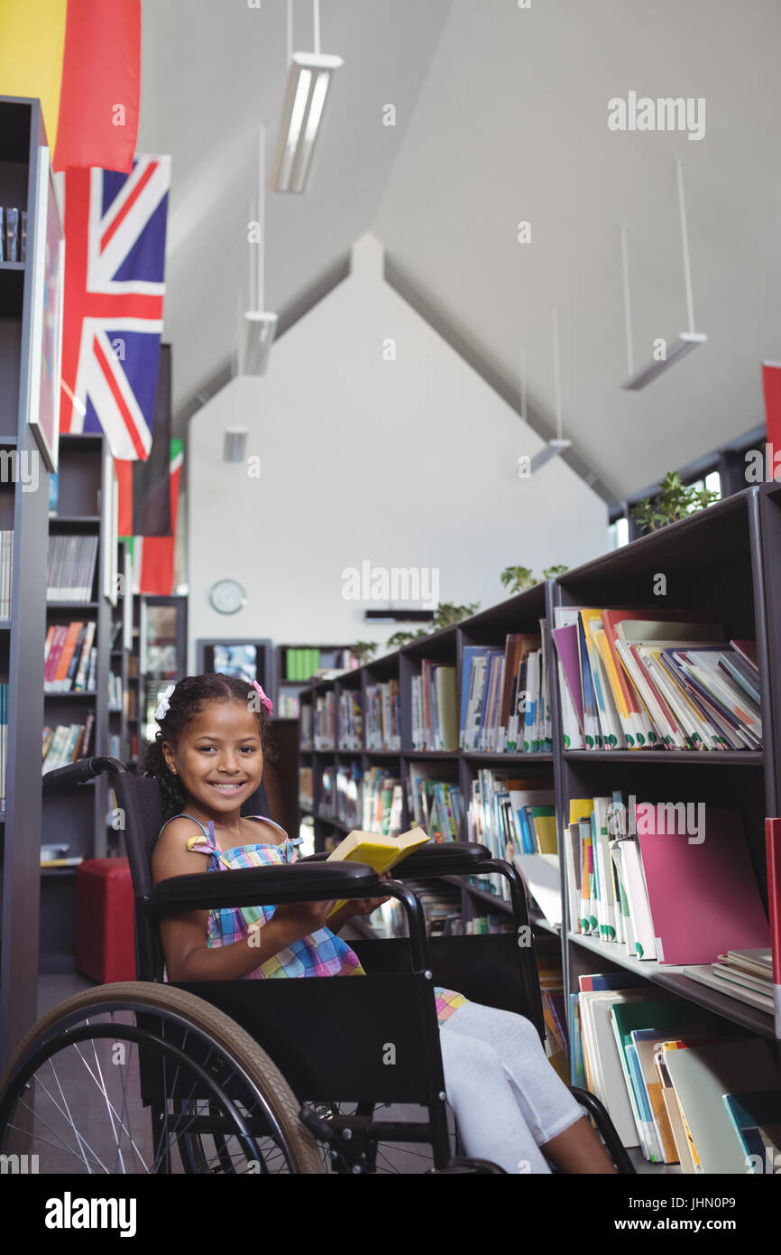 Portrait of smiling girl with book sitting on wheelchair by shelves in ...