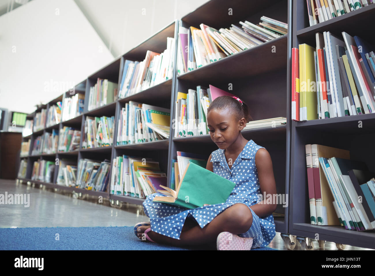 Girl reading book while sitting by shelf in library Stock Photo - Alamy
