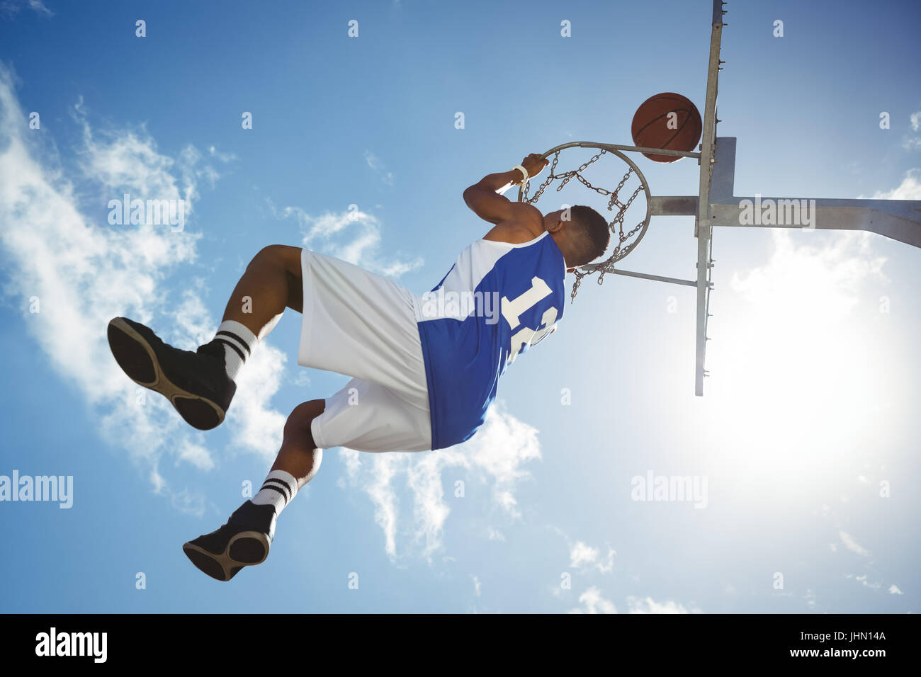 Low angle view of male teenager hanging on basketball hoop against blue ...