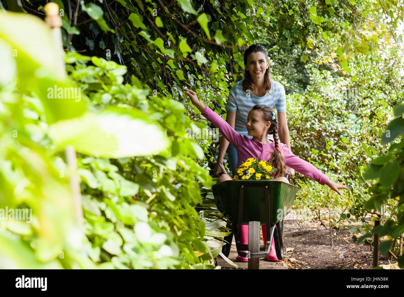 Smiling woman pushing girl sitting with outstretched in wheelbarrow on ...