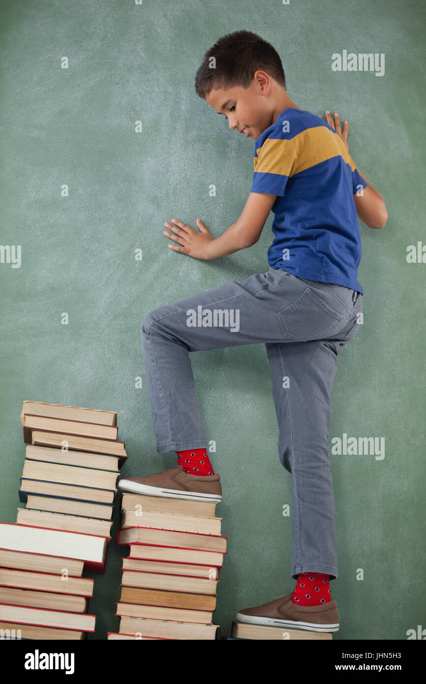 Schoolboy climbing steps of books stack against chalkboard Stock Photo ...