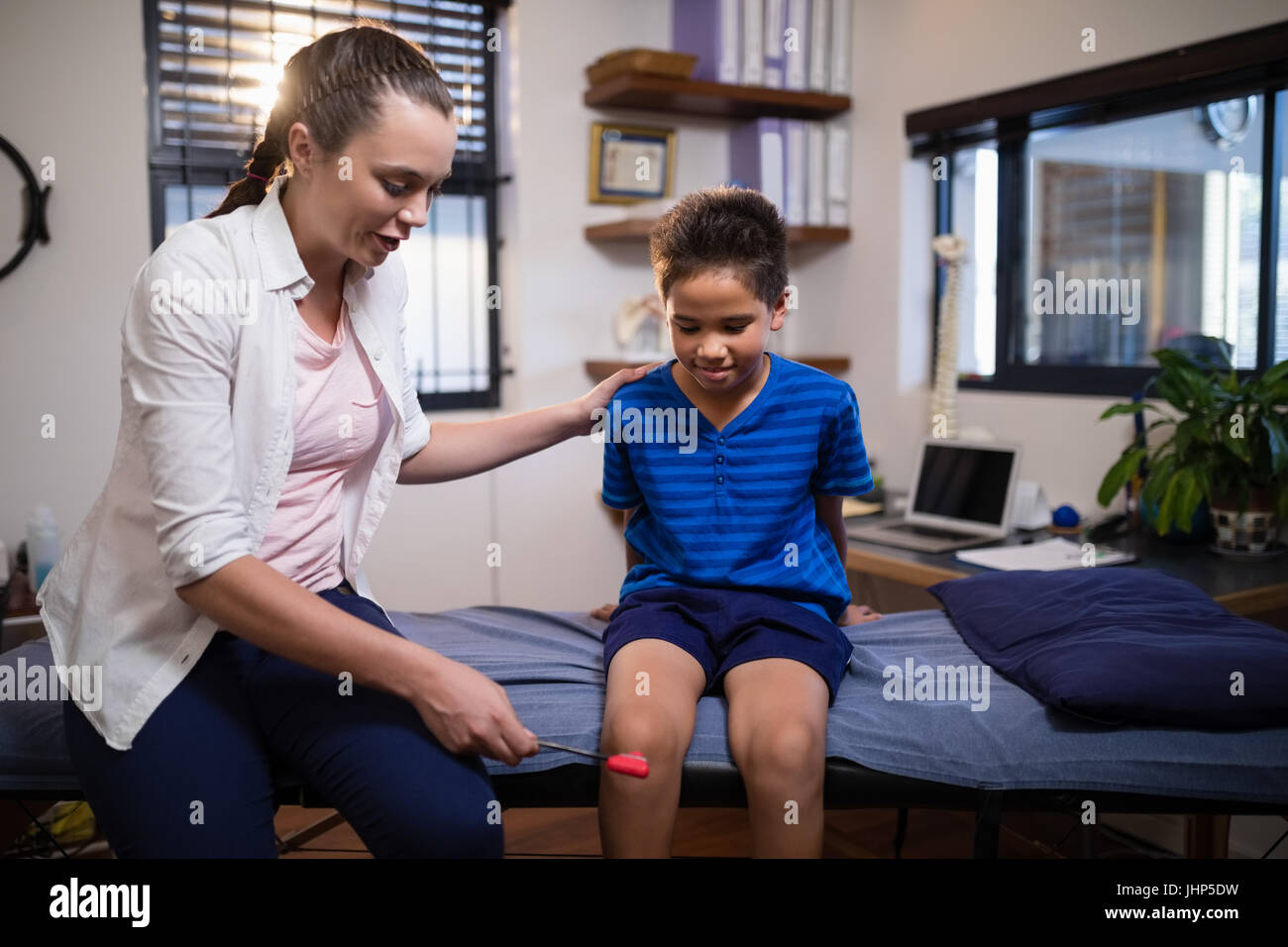 Young female therapist examining boy with reflex hammer on knee while ...