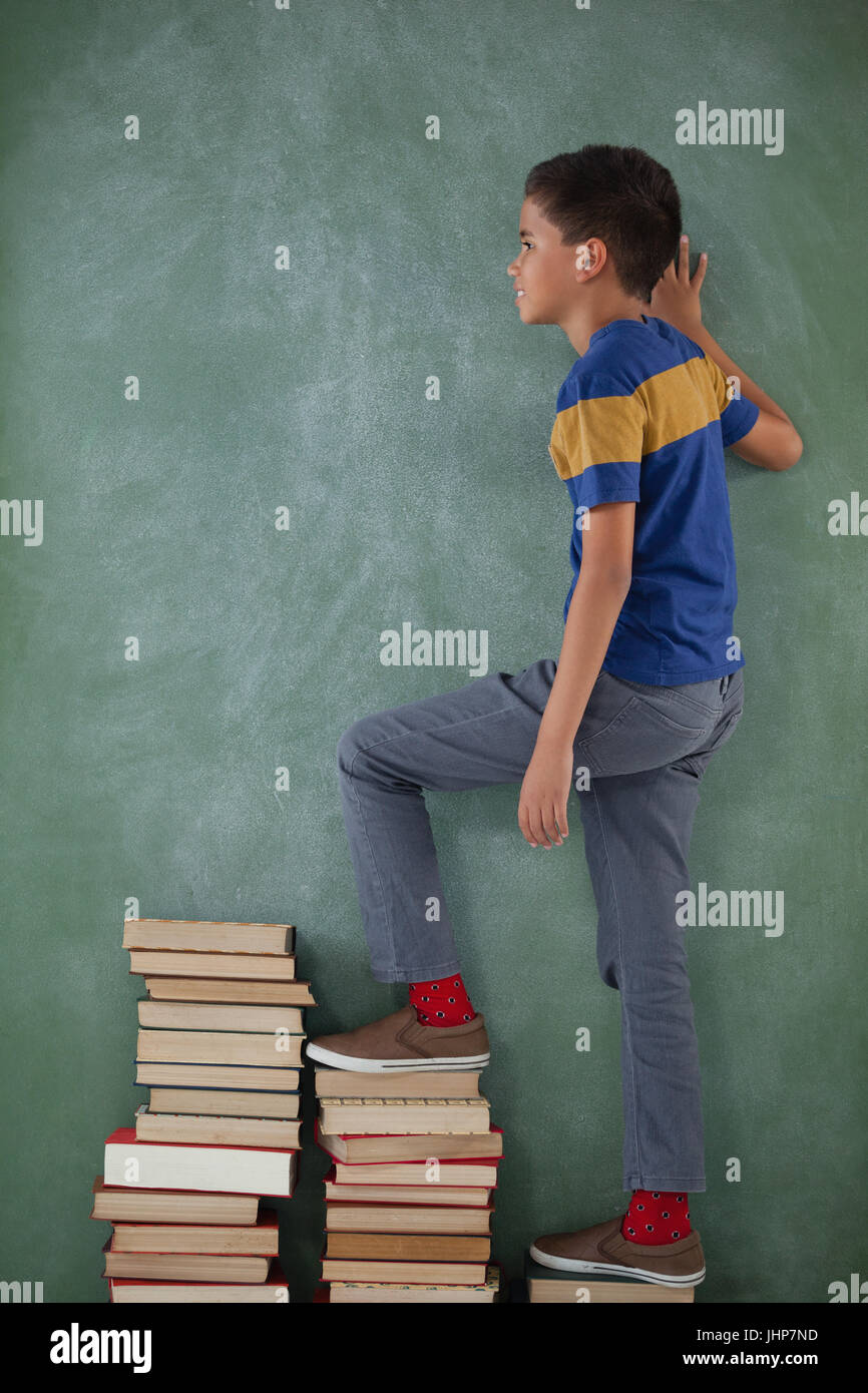 Schoolboy climbing steps of books stack against chalkboard Stock Photo ...