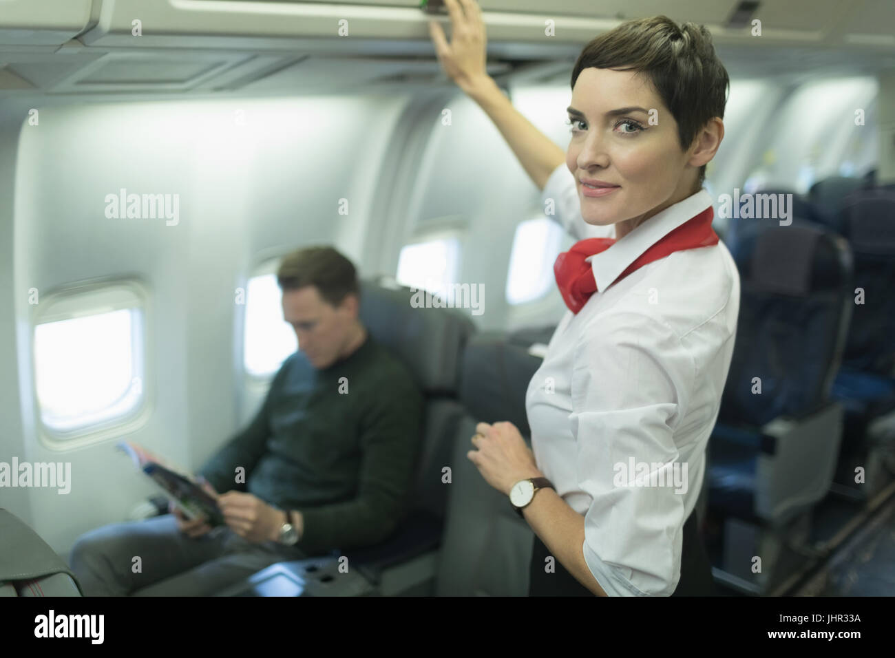 Confident air hostess smiling at camera while passenger reading ...