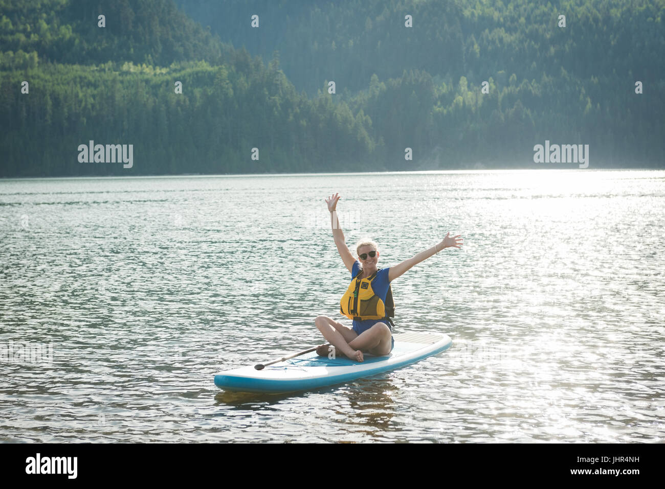 Excited young woman sitting with arms raised on paddleboard in lake ...